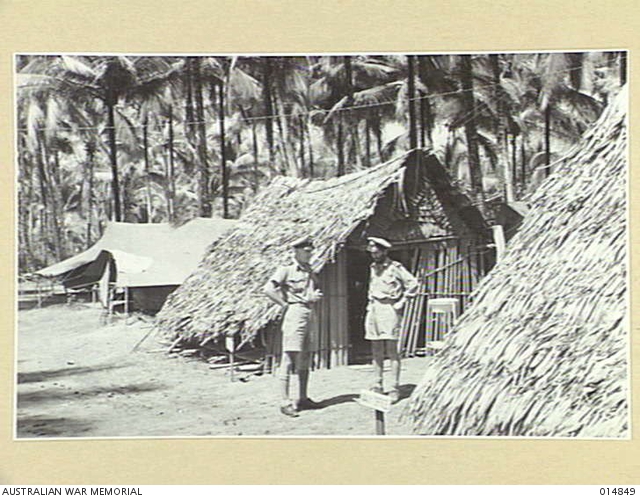 Two naval officers standing between two locally made huts in use at a ...