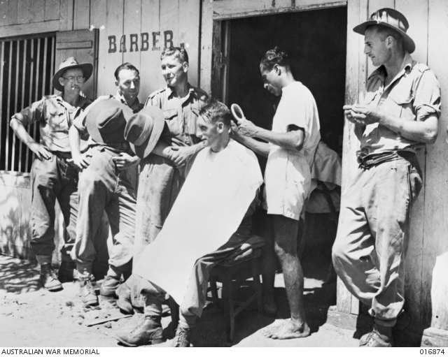 Dutch New Guinea. Soldiers line-up against the building for a haircut ...