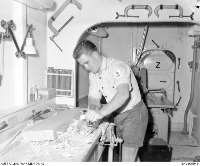A leading hand, possibly a shipwright, planing a piece of wood in a ...